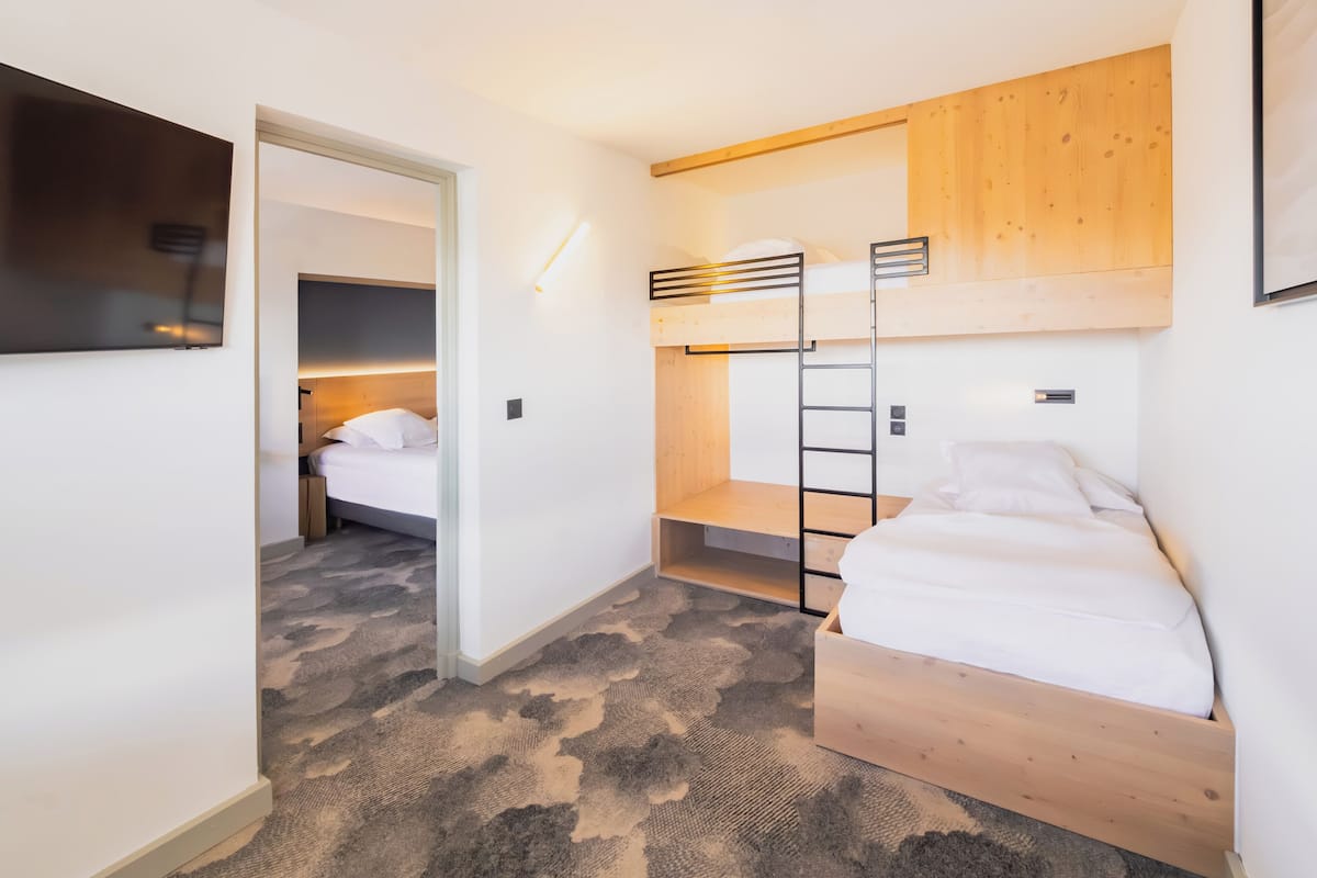 Quarto de hotel pequeno com beliches, televisão de tela plana na parede e piso coberto por carpete.