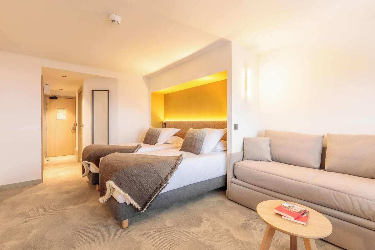 Quarto de hotel com duas camas, um sofá, uma mesinha e um corredor que leva a uma porta.