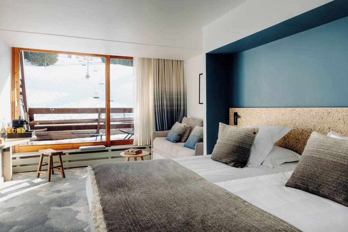 Quarto de hotel moderno com uma cama grande, um sofá, uma mesa pequena e vista para uma pista de esqui através de uma janela ampla.
