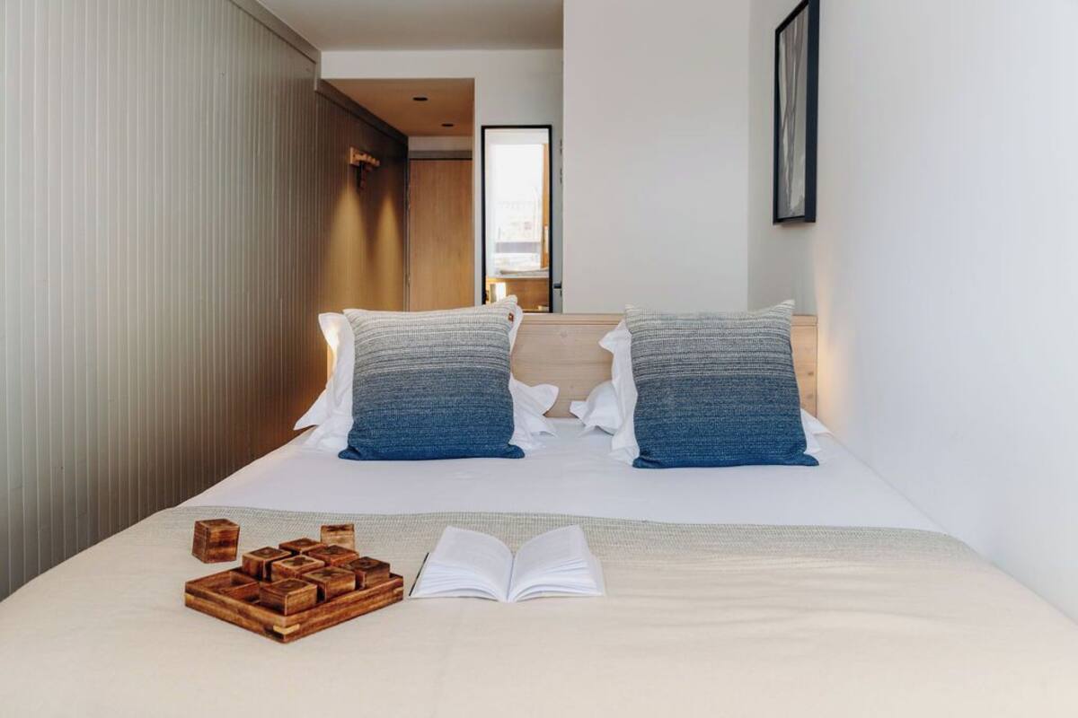 Cama bem arrumada com duas almofadas, bandeja de madeira com blocos e um livro aberto sobre a cama do quarto de hotel.