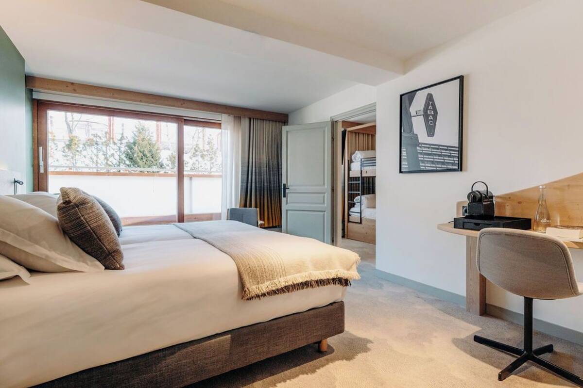Quarto com cama, escrivaninha e cadeira. Há uma janela com cortinas, uma porta que dá para outro cômodo e um quadro na parede.