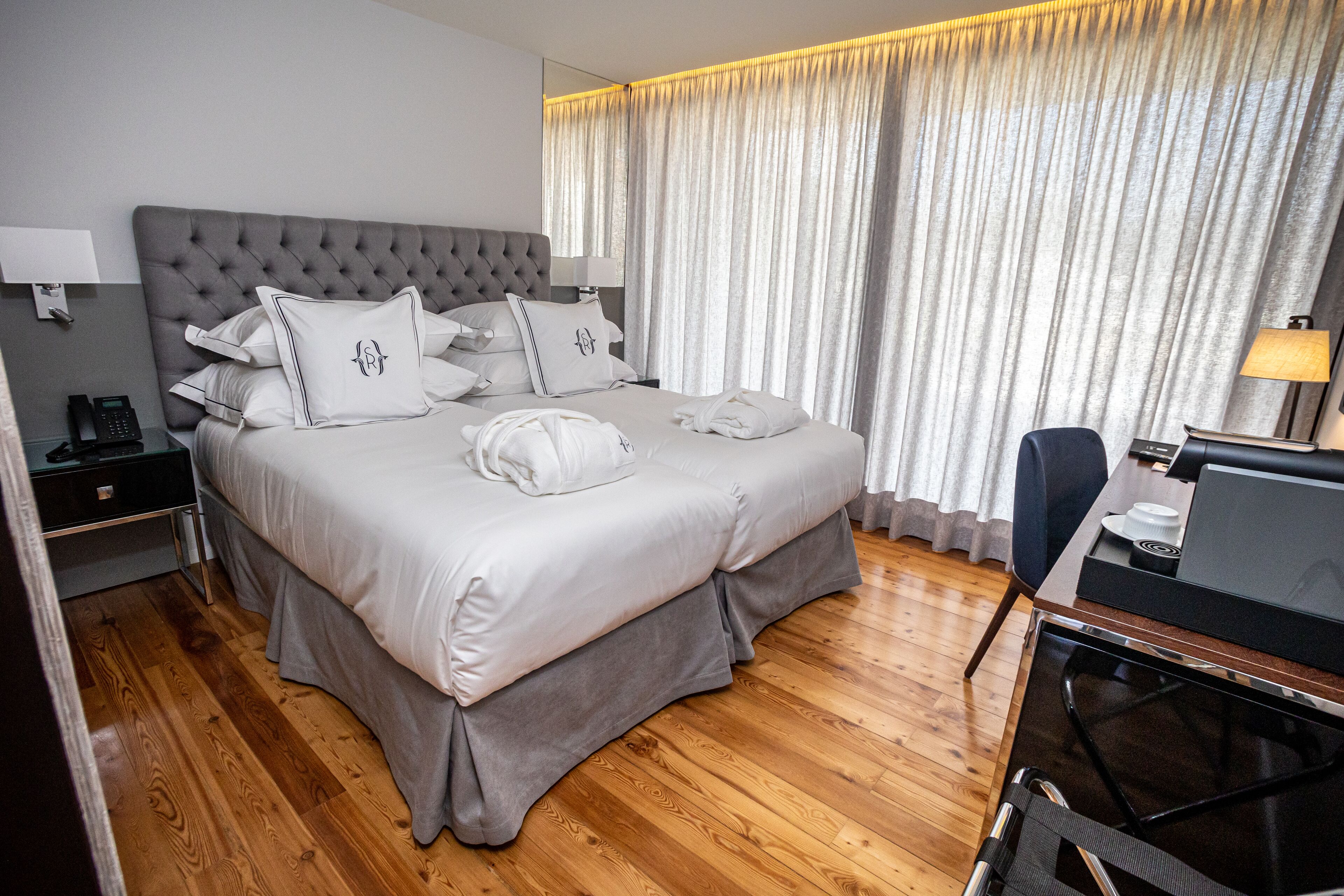 Quarto de hotel com uma cama grande, cabeceira estofada cinza, roupa de cama branca e uma escrivaninha com abajur.