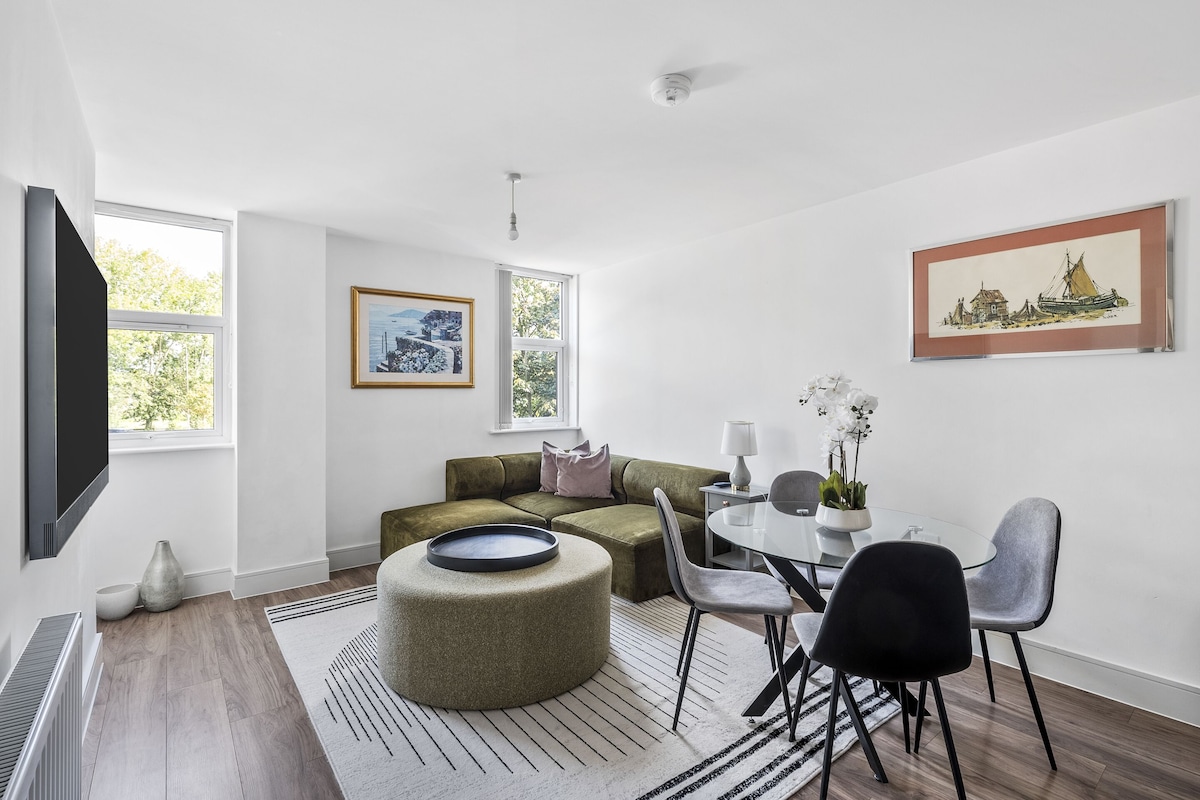 Uma sala de estar moderna com um sofá modular, um banquinho redondo, uma mesa de centro de vidro e uma área de jantar com cadeiras e uma luminária. Há duas janelas com vista para vegetação, e obras de arte estão expostas nas paredes.