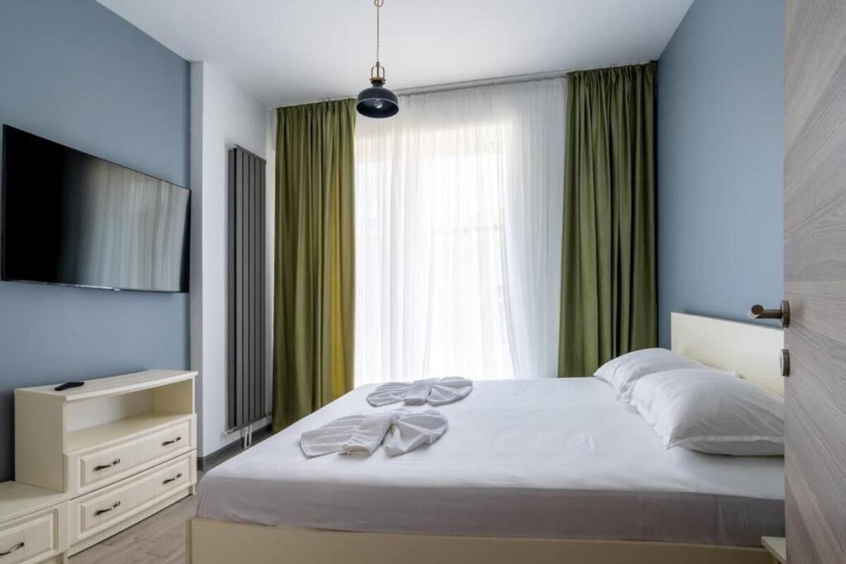 Quarto de hotel com cama, televisão fixada na parede, cómoda e janela com cortinas.