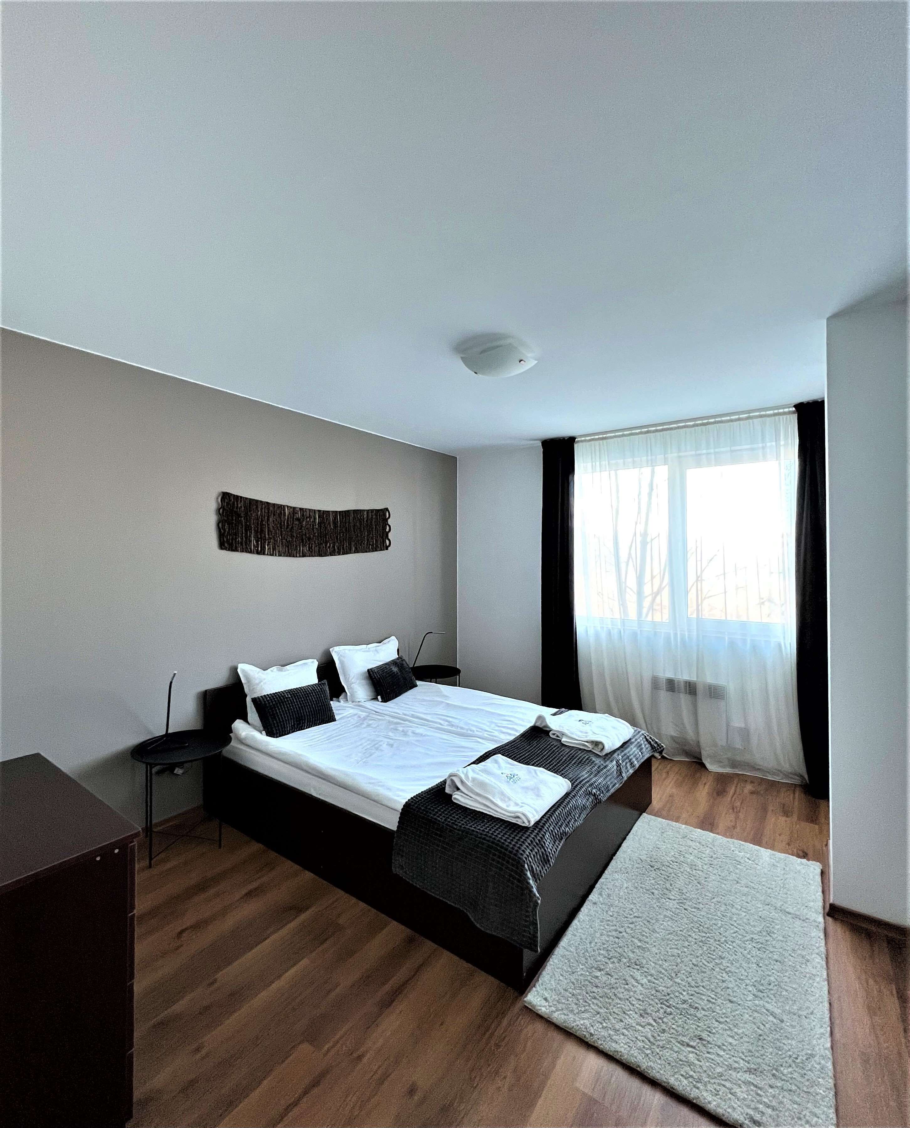 Quarto moderno com cama grande, mesa de cabeceira, escrivaninha e janela com cortinas.