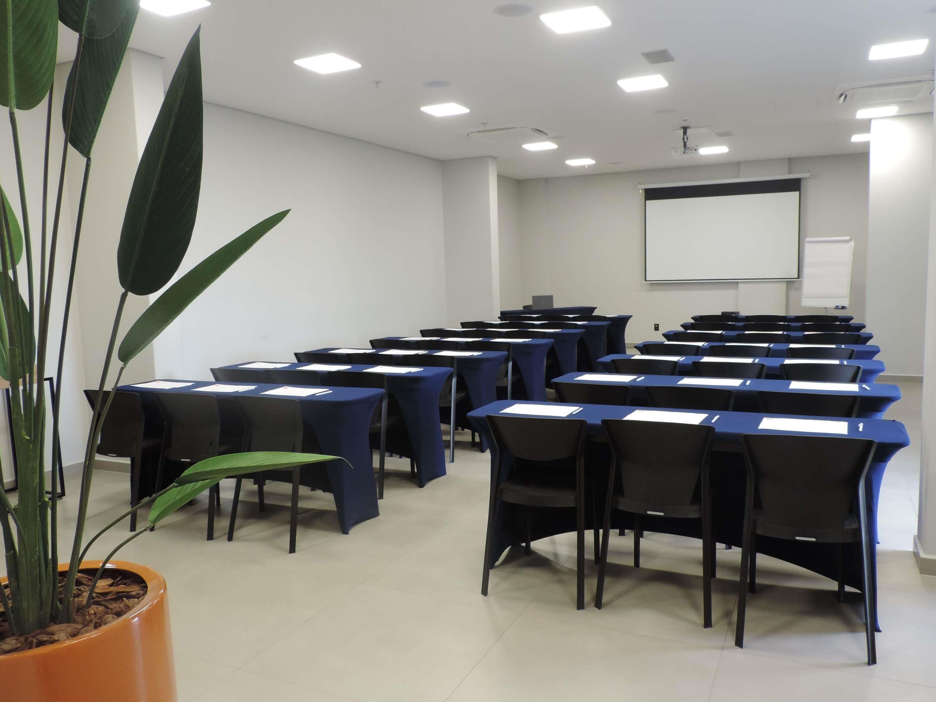 Uma sala de conferência com fileiras de mesas e cadeiras, uma tela de projetor e um vaso de planta.