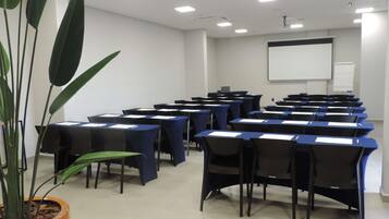 Uma sala de conferência com fileiras de mesas e cadeiras, uma tela de projetor e um vaso de planta.
