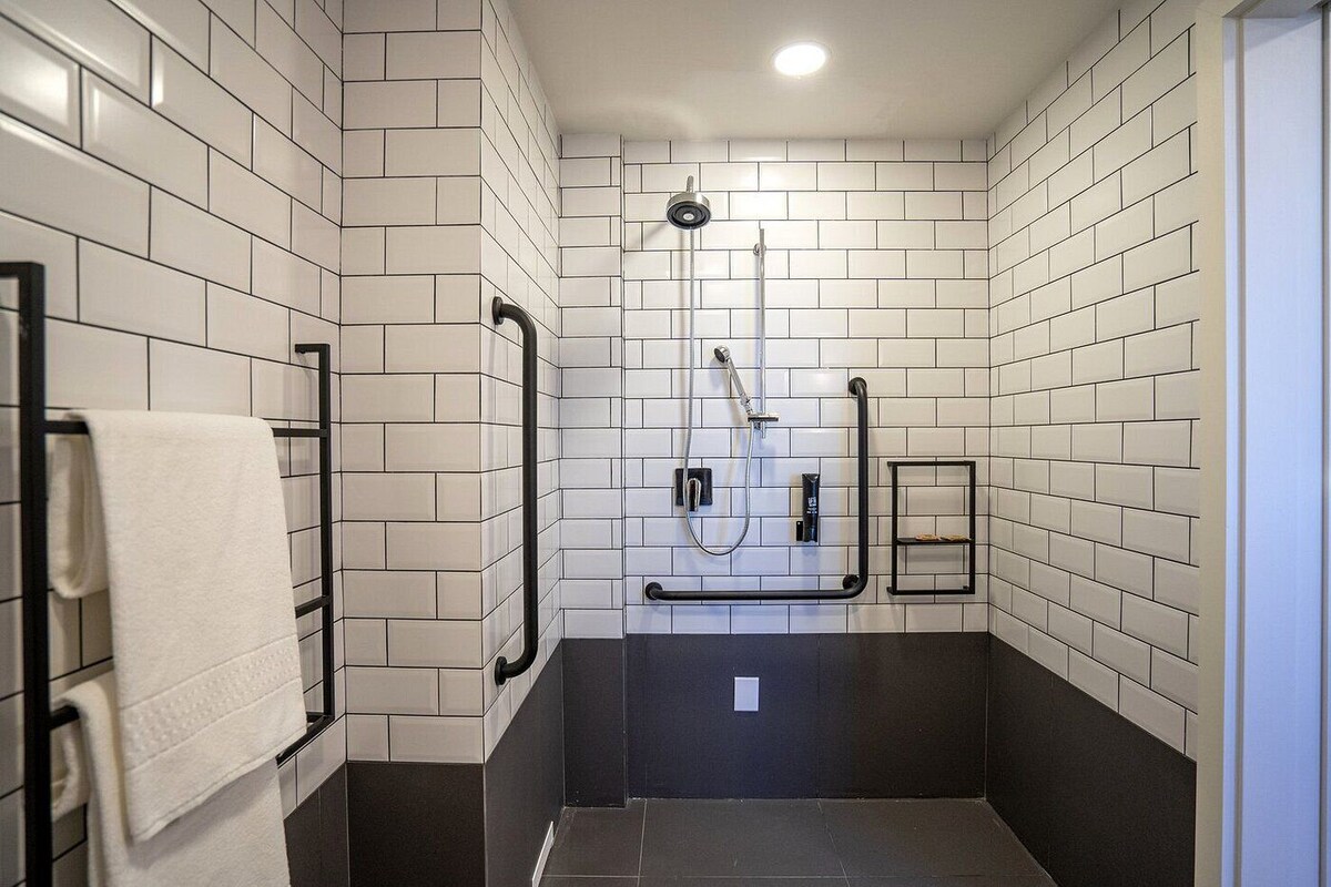 Casa de banho moderna com paredes em azulejos brancos e pretos, suporte de toalhas preto, chuveiro com chuveiro manual e um pequeno prateleira.