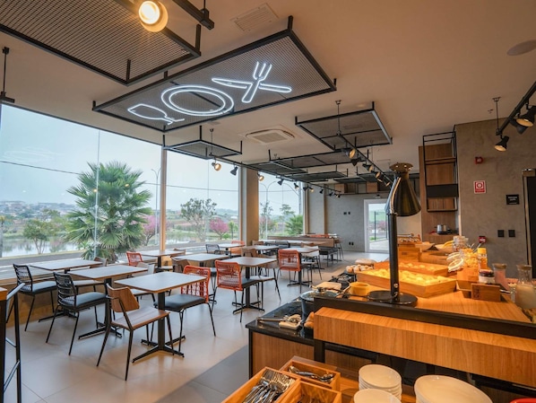 Restaurante moderno com vista para um lago, apresentando um letreiro de neon, mesas de madeira e uma área de buffet.