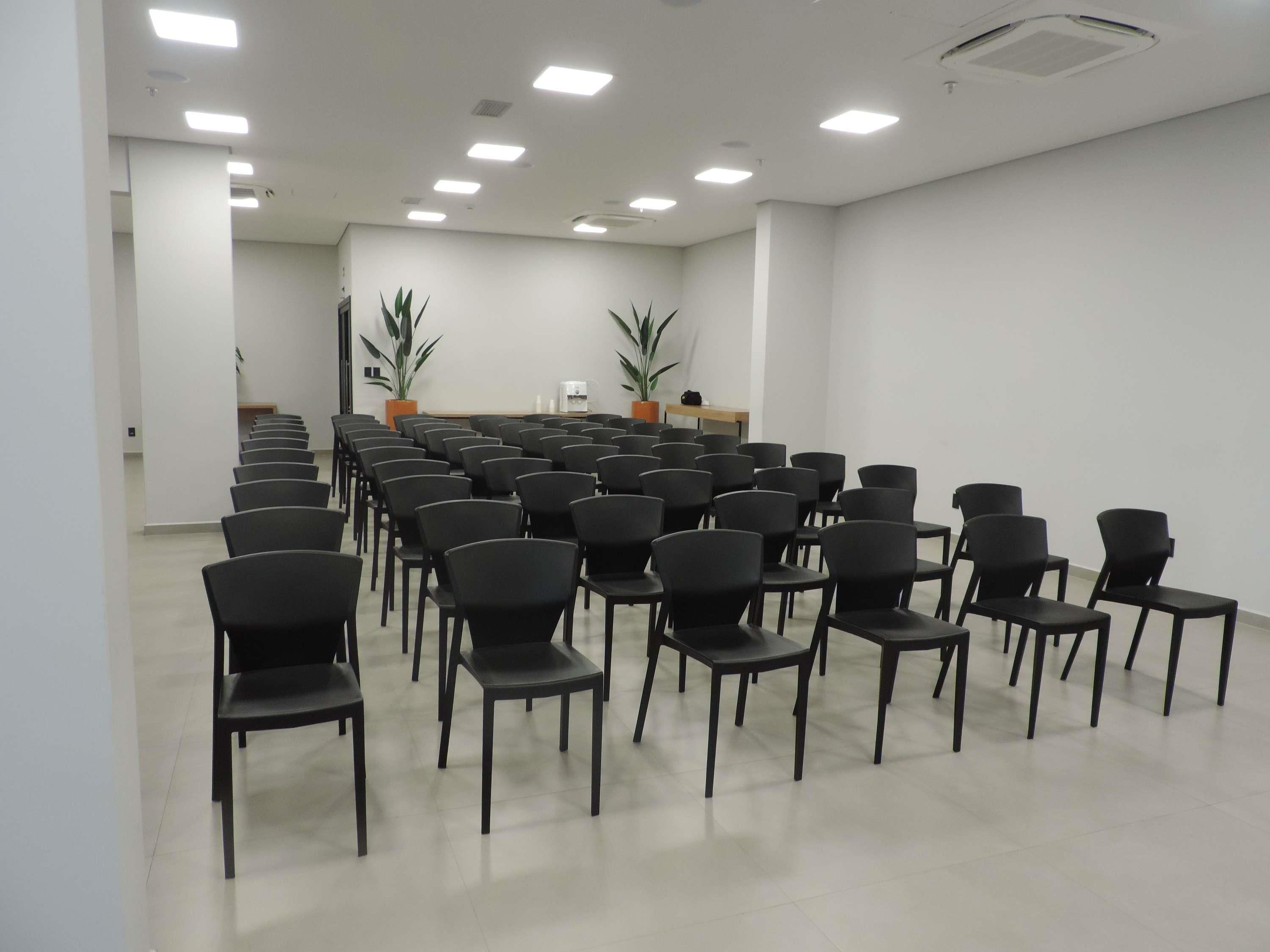 Uma sala de conferências com fileiras de cadeiras pretas, paredes brancas e luminárias no teto.