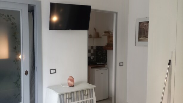 Um corredor com uma porta, uma televisão presa na parede, uma mesa pequena com um objeto decorativo e um frigorífico ao fundo.