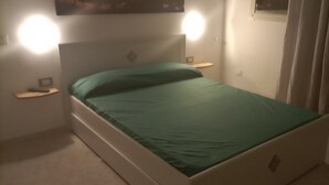 Uma cama de solteiro com colchão verde em quarto com paredes brancas. Há duas mesas de cabeceira, cada uma com uma luminária. Uma janela com cortinas é visível à direita.