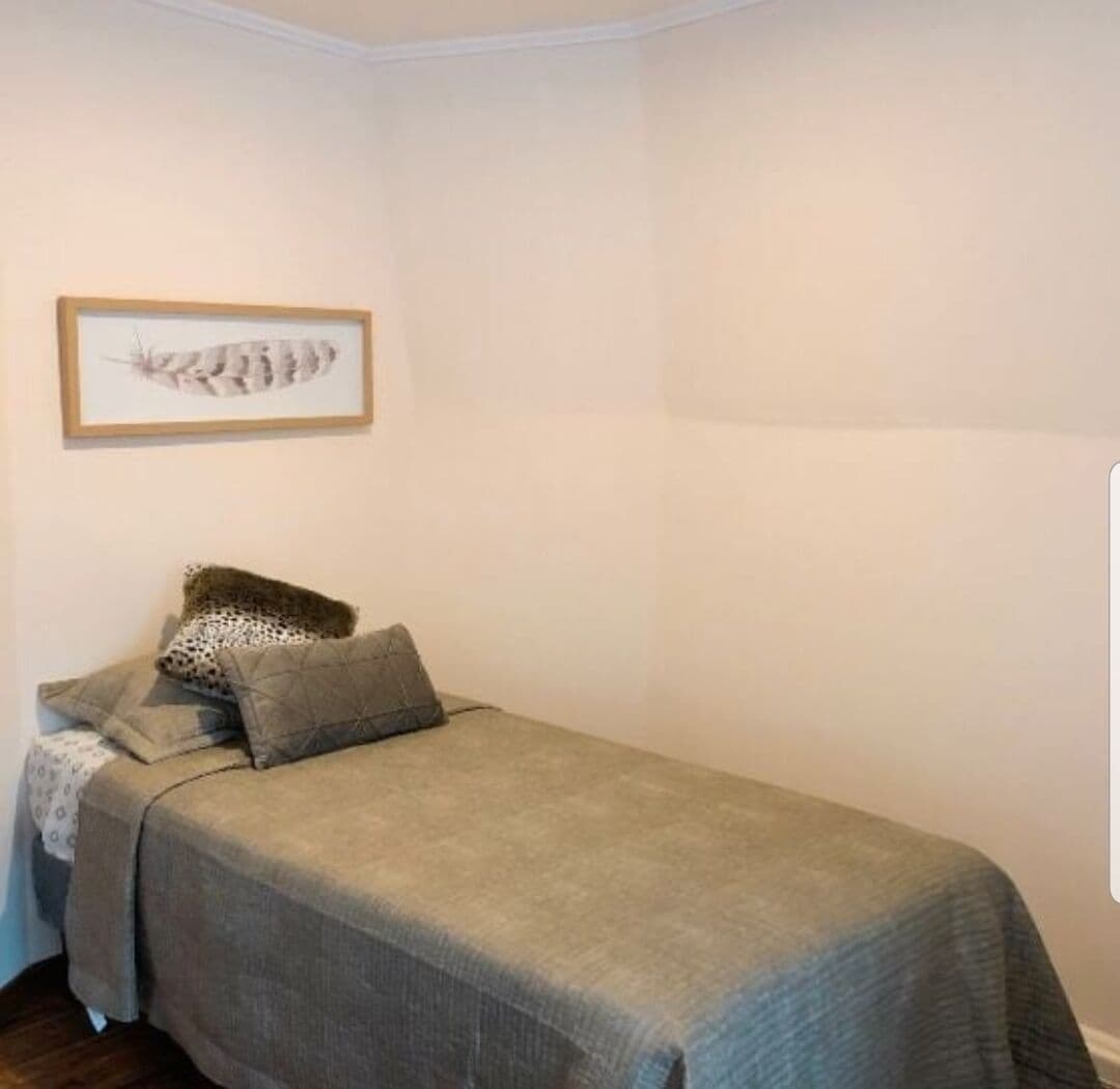 Uma cama pequena e bem arrumada, com um edredom cinzento e duas almofadas, uma com padrão de leopardo. Um quadro emoldurado está pendurado na parede acima da cama.