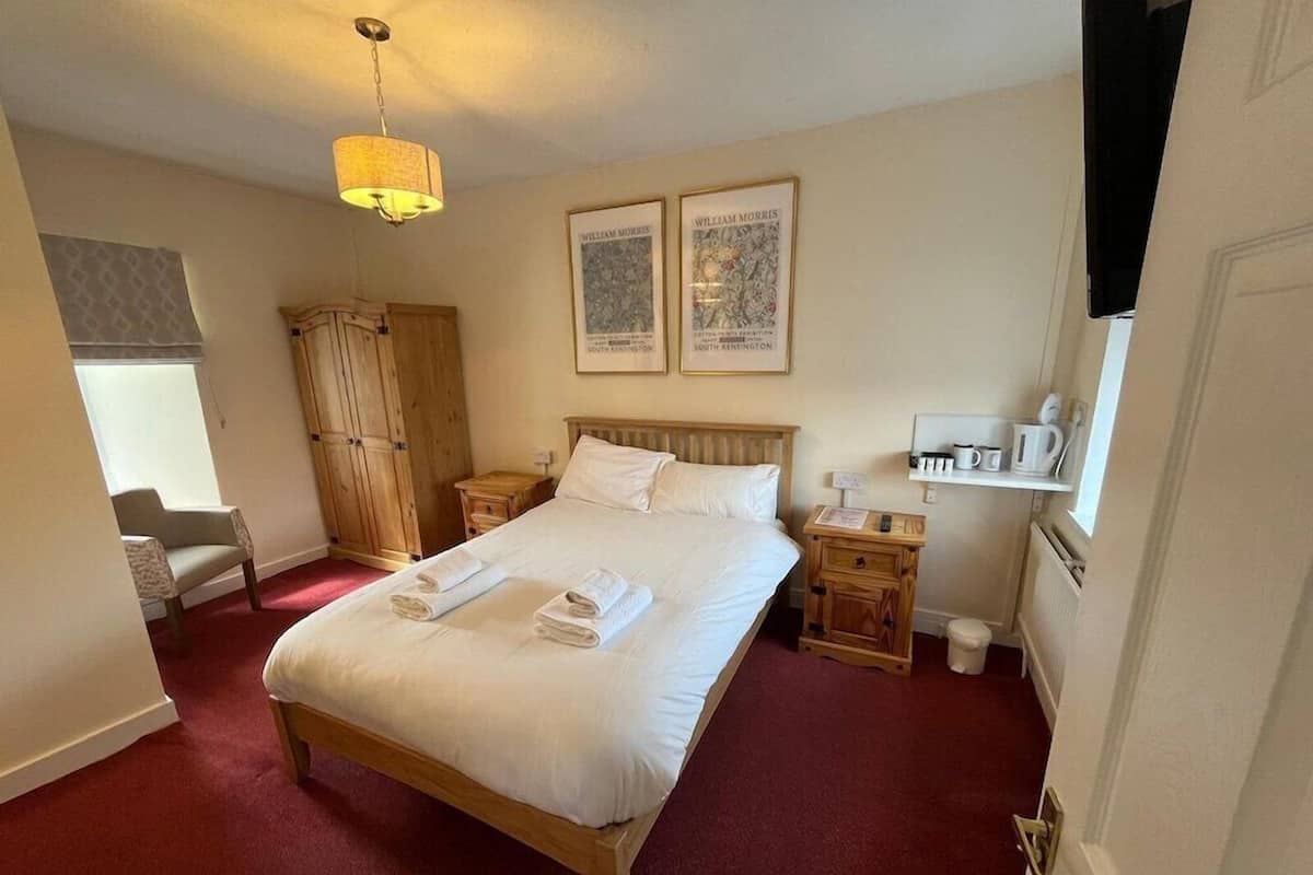 Quarto de hotel com uma cama grande, guarda-roupa de madeira, mesas de cabeceira, uma cadeira e duas imagens emolduradas na parede.
