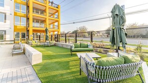 Um pátio moderno ao ar livre com gramado verde, mobiliário de vime e um edifício amarelo ao fundo.