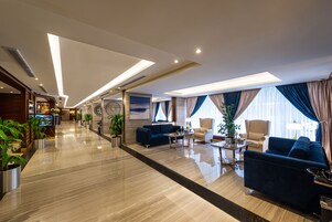 Um hall de hotel moderno com piso de mármore polido, um sofá azul, uma poltrona bege e uma mesa de centro. Há plantas em vasos e uma grande janela com cortinas.