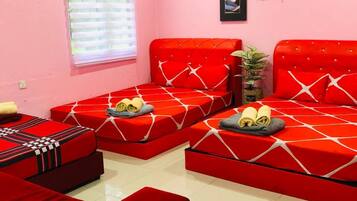 Quarto com parede rosa, dois sofás vermelhos, uma mesa pequena com toalhas e uma janela com persianas.