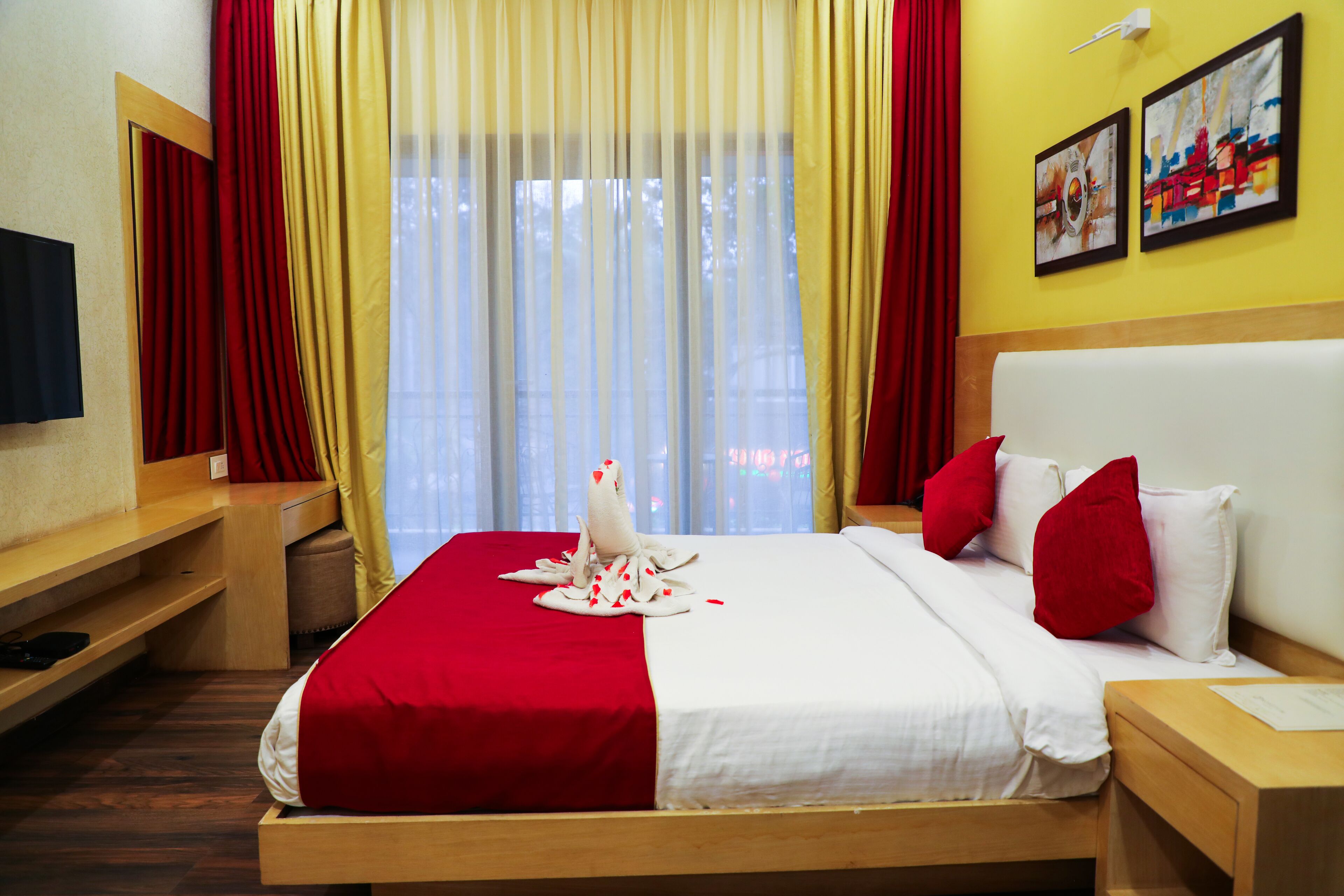 Quarto de hotel com cama, roupa de cama vermelha e branca, cabeceira de madeira, mesa de cabeceira de madeira, cortina vermelha e branca, janela com cortinas finas e duas obras de arte emolduradas na parede.