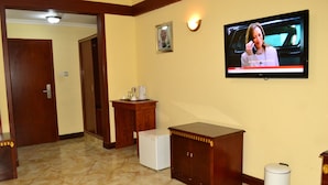 Quarto de hotel com televisão presa na parede, um frigorífico branco pequeno, um armário de madeira e um cesto de lixo.