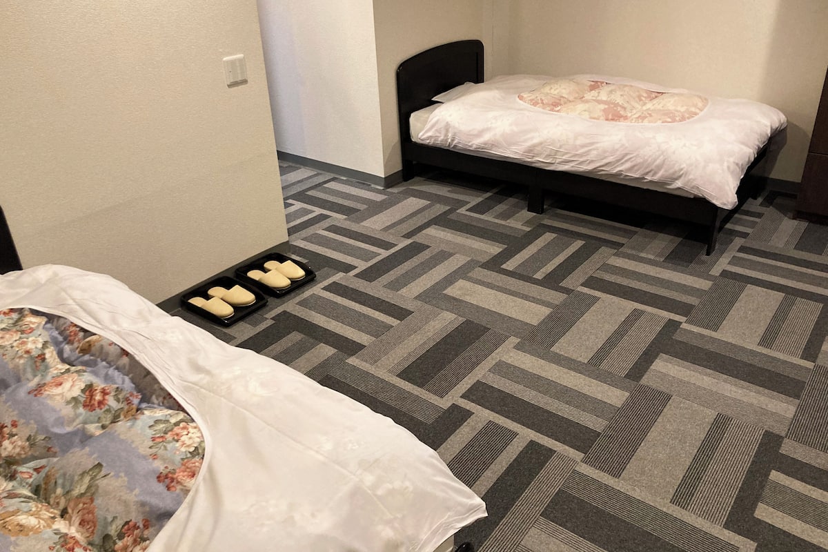 Quarto de hotel com duas camas, um tapete estampado e um suporte com chinelos.