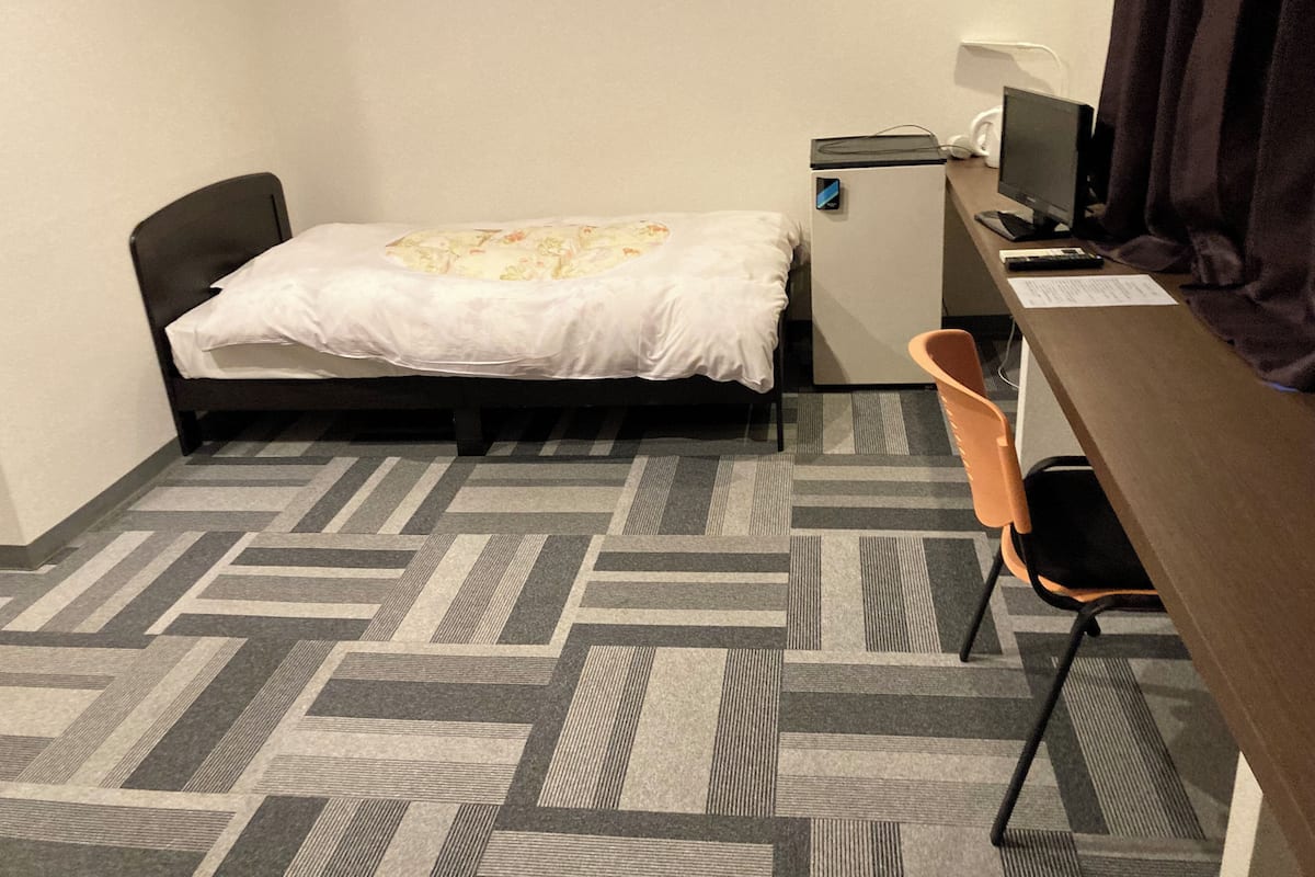 Um quarto de hotel pequeno com uma cama, uma escrivaninha, uma cadeira, um refrigerador e uma televisão.