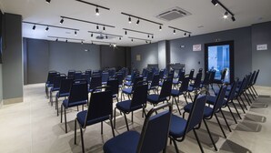Uma sala de conferências com fileiras de cadeiras azuis, uma tela de projetor e luminárias no teto.