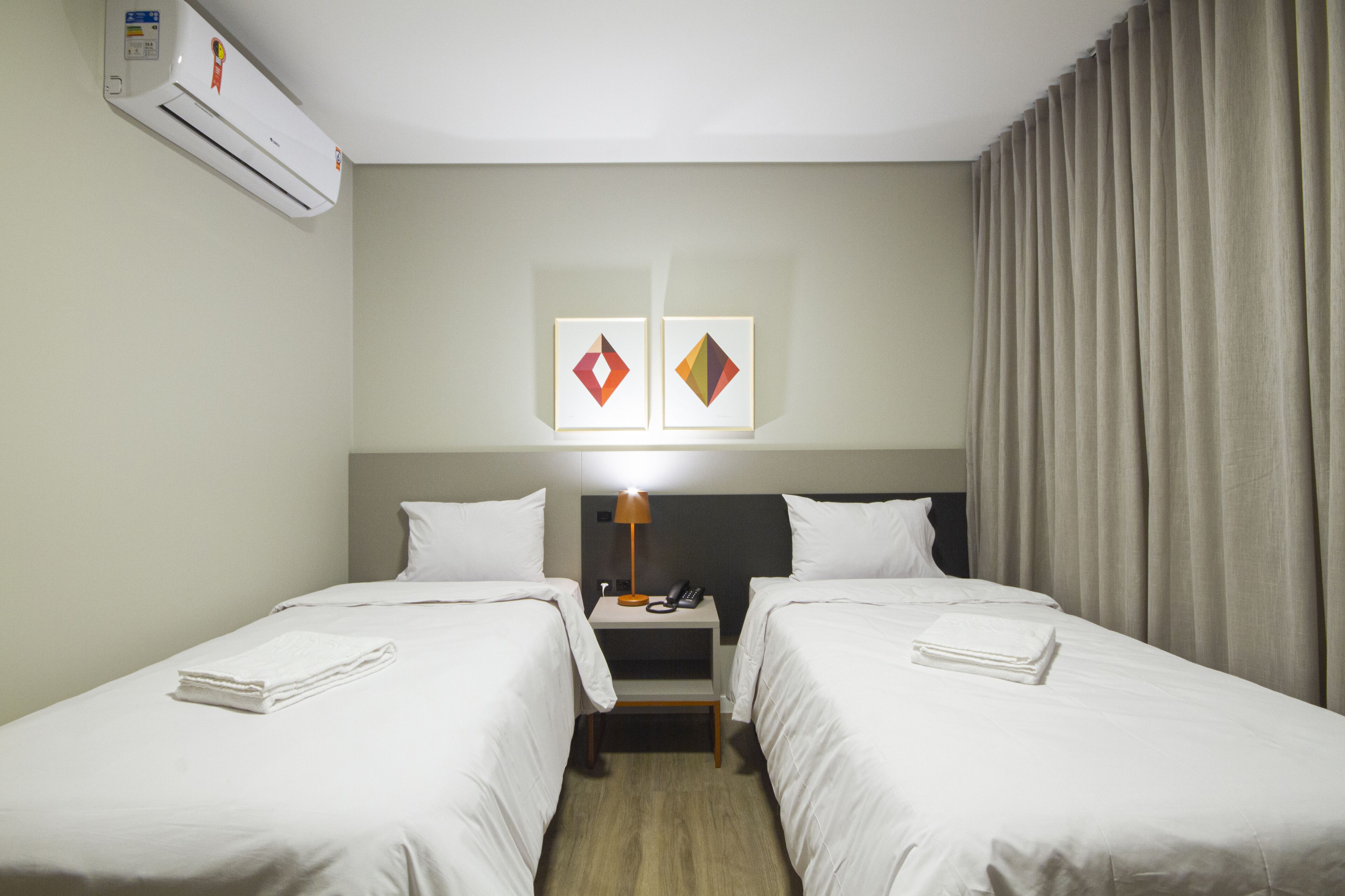 Quarto de hotel com duas camas, uma mesa de cabeceira com luminária, telefone e aparelho de ar condicionado.