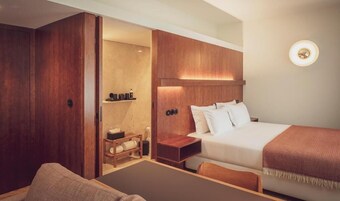 Quarto de hotel moderno com guarda-roupa de madeira, cama com lençóis brancos, mesa de cabeceira com abajur e uma pequena prateleira com artigos de higiene pessoal.
