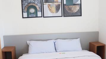 Cama bem arrumada com edredom branco e duas almofadas, com mesas de cabeceira de madeira em ambos os lados. Acima da cama, três obras de arte emolduradas penduradas na parede.