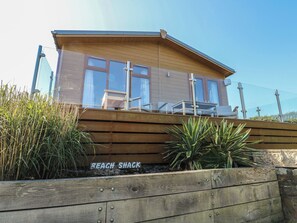 Um edifício de madeira com varanda, identificado como 'Beach Shack', e cerca de madeira com plantas à frente.