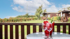 Mesa de vidro com um vaso vermelho e uma taça de vinho numa varanda com vista para uma área gramada e cabanas de madeira.