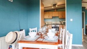 Uma sala de jantar com mesa de madeira posta para seis pessoas, cadeiras brancas e cozinha visível ao fundo.