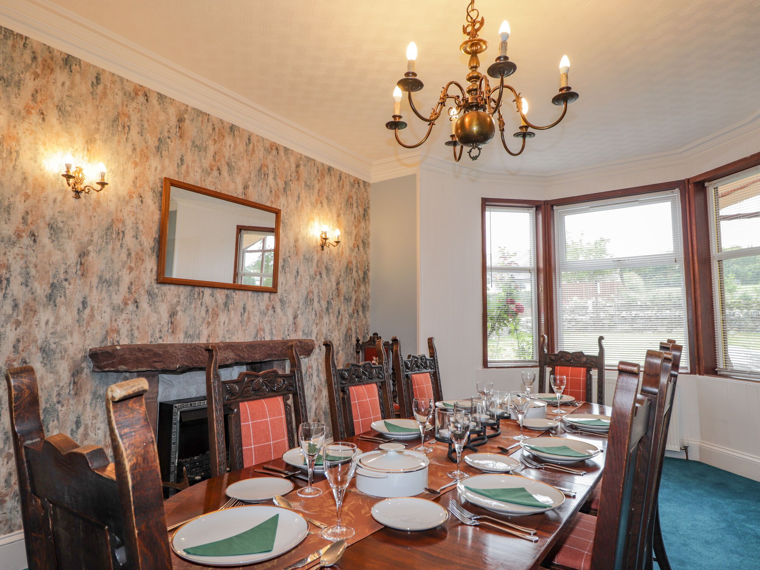 Uma sala de jantar com uma longa mesa de madeira posta para uma refeição, lareira e um lustre.