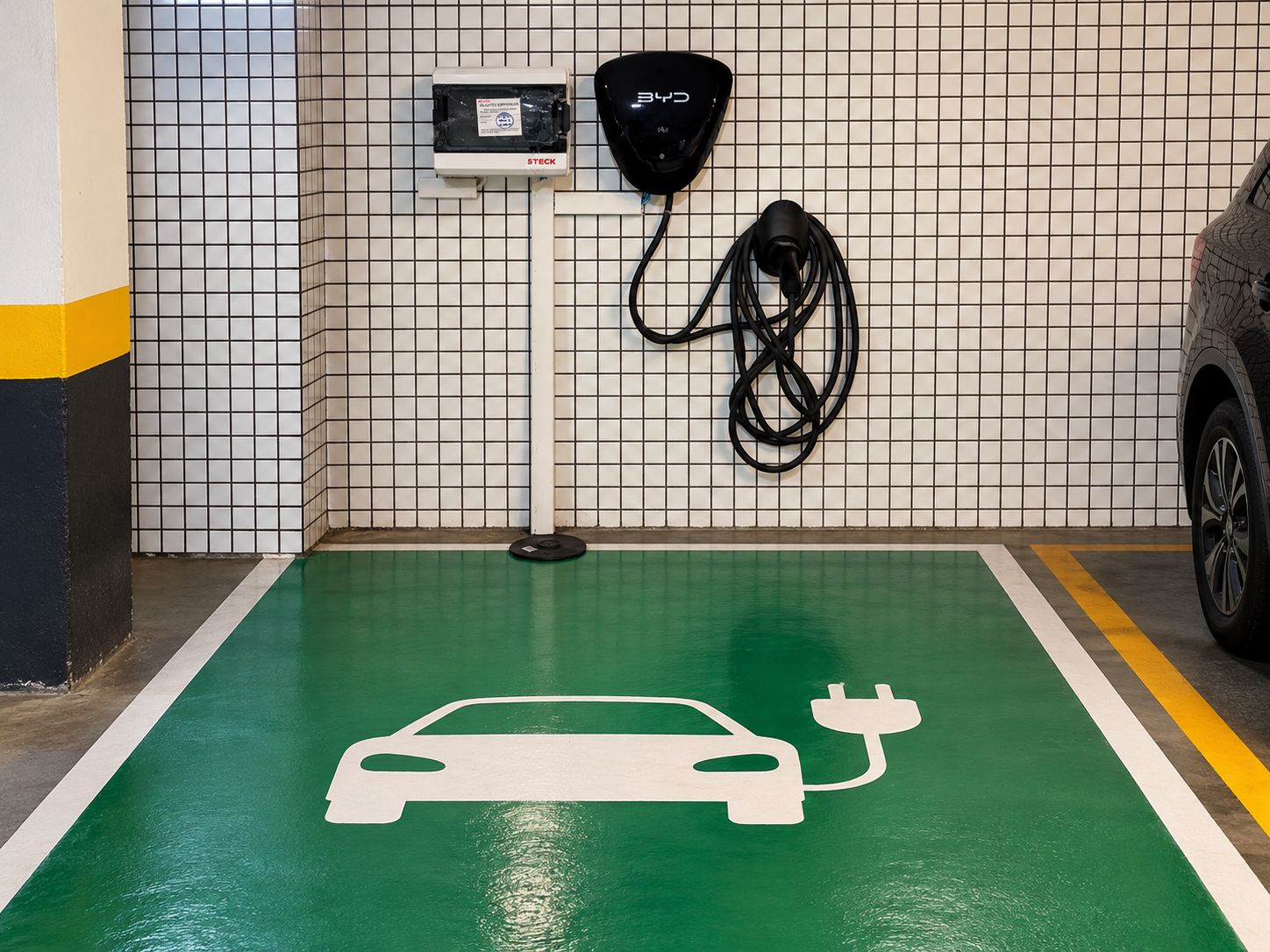 Vaga de estacionamento com piso verde demarcada para veículos elétricos, uma estação de carregamento e um carro estacionado à direita.