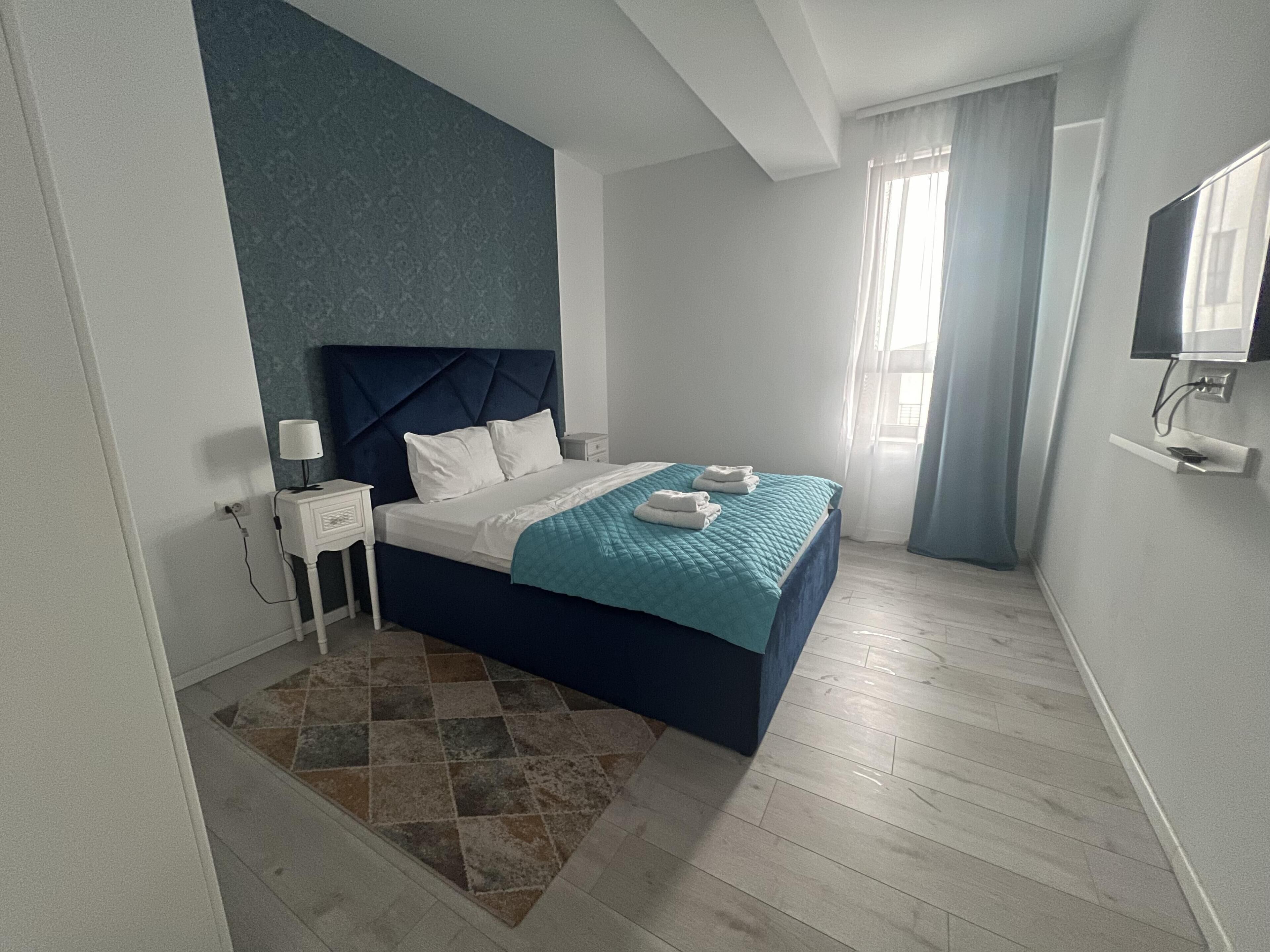 Quarto moderno com uma cama grande, mesa de cabeceira, televisão montada na parede e janela com cortinas.