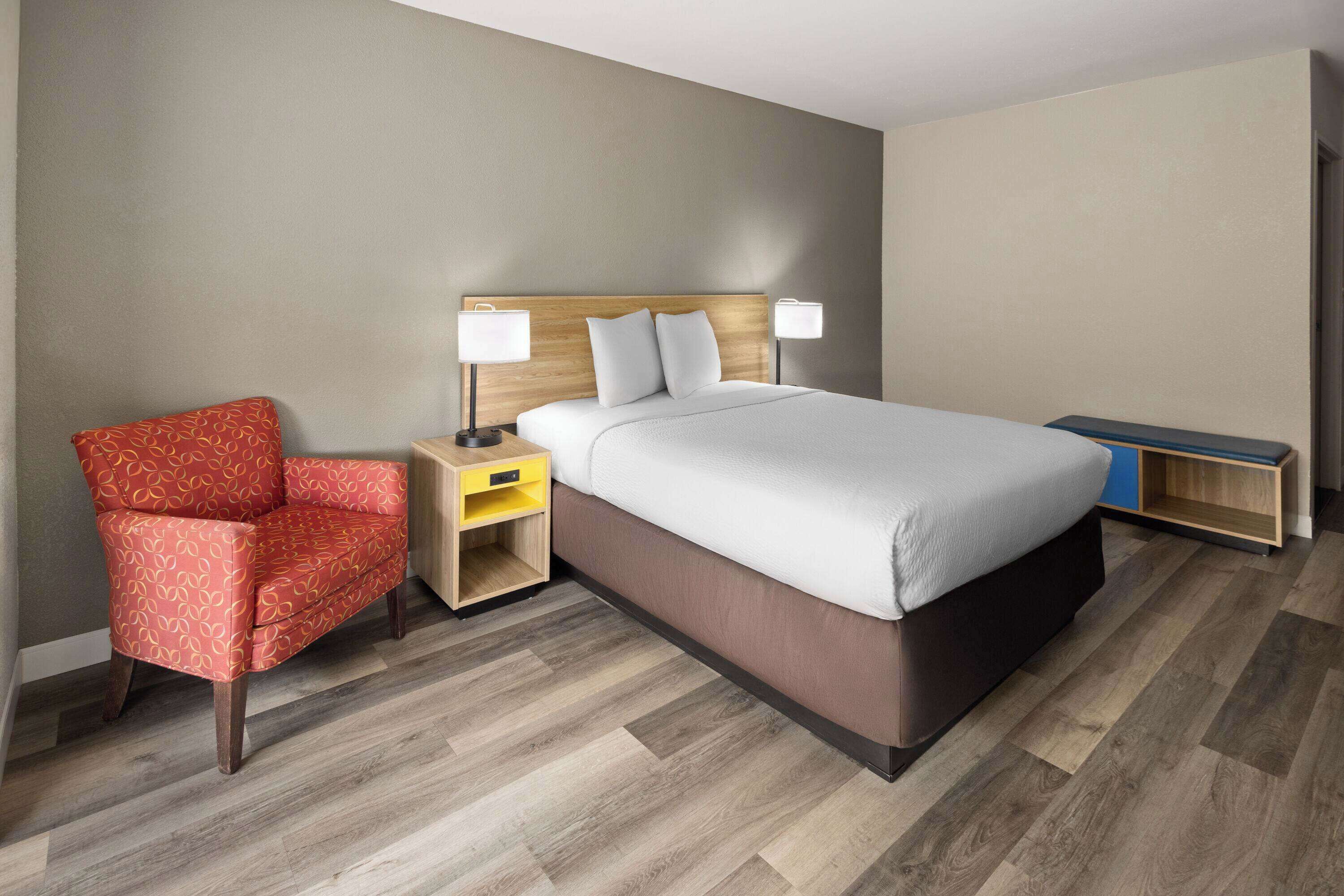 Quarto de hotel com uma cama grande, uma mesa de cabeceira com abajur, uma cadeira e um pequeno armário de madeira.