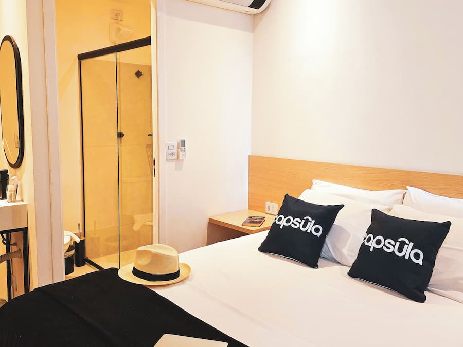 Quarto de hotel com uma cama, duas almofadas pretas com o texto 'capsula', um laptop, um chapéu e um espelho na parede.
