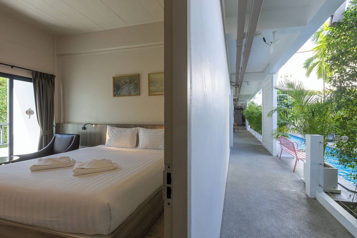 Quarto de hotel com cama, uma cadeira e vista para piscina e vegetação.