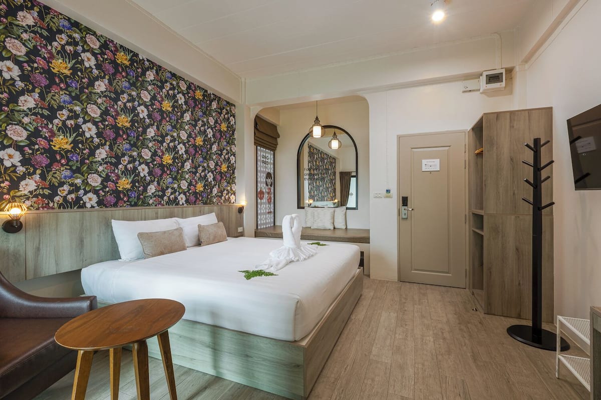 Quarto de hotel com uma cama grande, cabeceira de madeira, papel de parede floral, espelho, guarda-roupa de madeira e um cabideiro.
