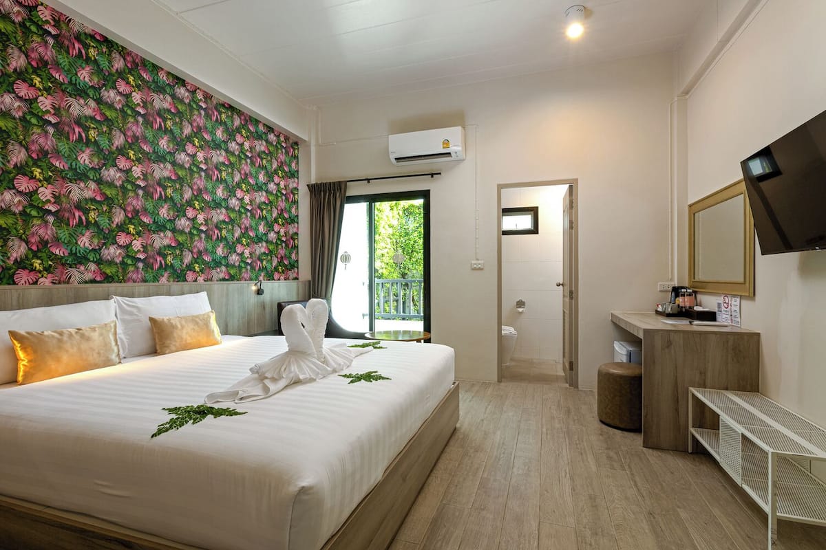 Quarto de hotel com uma cama grande, cabeceira de madeira, televisão fixada na parede, escrivaninha com espelho e varanda com vista para o verde.