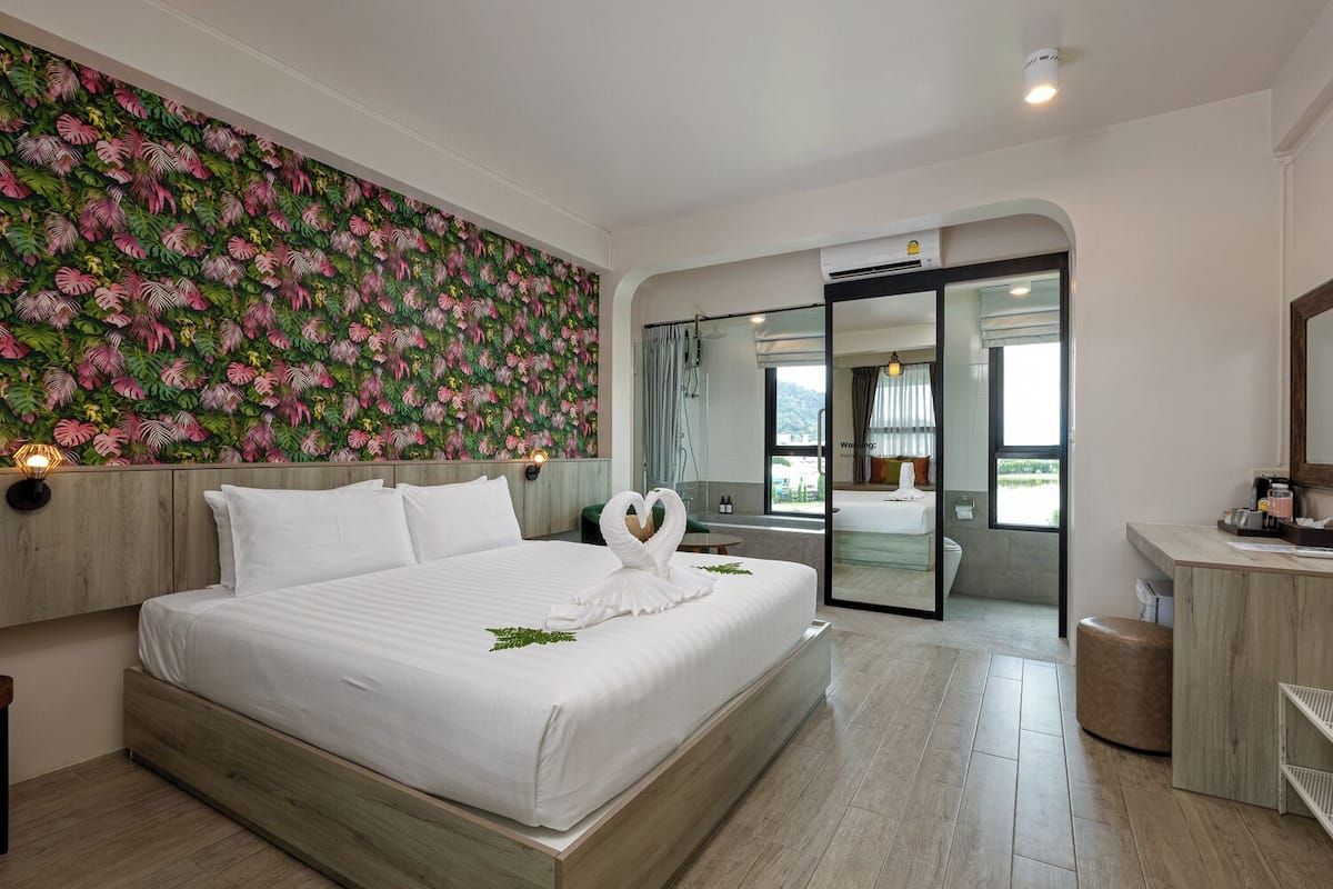 Quarto de hotel com uma cama grande, cabeceira de madeira, parede floral, banheiro com porta de vidro e escrivaninha com espelho.