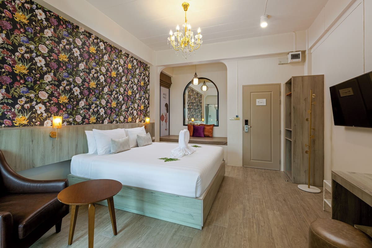 Quarto de hotel com uma cama grande, cabeceira de madeira, uma mesinha, um sofá, uma televisão e papel de parede floral.