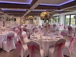 Uma sala de banquetes com mesas redondas, cadeiras brancas com fitas cor-de-rosa e centros de mesa florais.