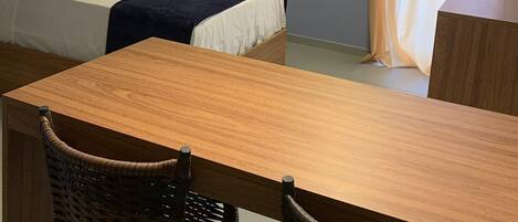 Quarto de hotel com cama de madeira, escrivaninha de madeira, duas cadeiras de vime, luminária pendente e uma janela com cortinas.
