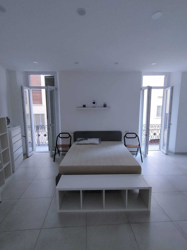 Quarto com uma cama grande, duas cadeiras, um pequeno estante e vista para a varanda.