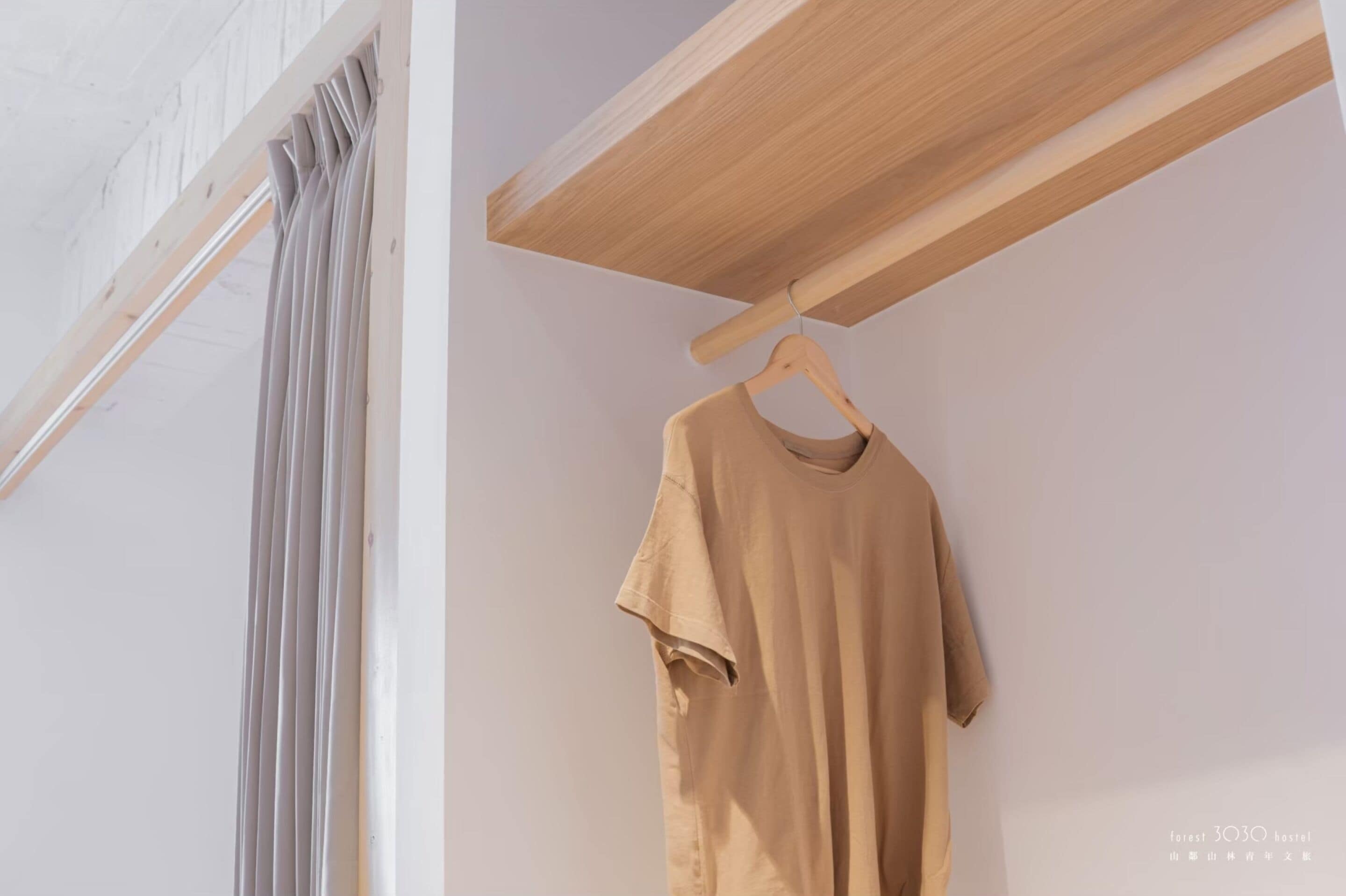 Quarto com uma viga de madeira no teto, uma t-shirt bege pendurada e uma janela com persianas brancas.