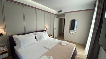 Quarto de hotel com uma cama grande, mesa de cabeceira com abajur, espelho na parede e porta para outro quarto.