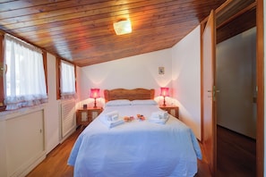 Quarto com teto de madeira, cama com roupa de cama branca, duas luminárias de cabeceira e um radiador.