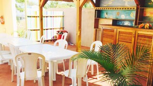 Uma área de refeições ao ar livre com cadeiras e mesas de plástico branco, uma pérgola de madeira e um mural na parede.