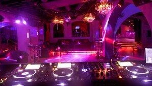 Um espaço DJ com várias plataformas e misturadores, rodeado por uma área de bar com assentos e uma escadaria.